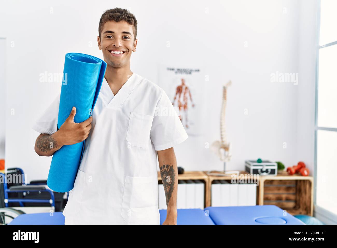 Young hispanic man working as physiotherapist holding yoga mat at ...