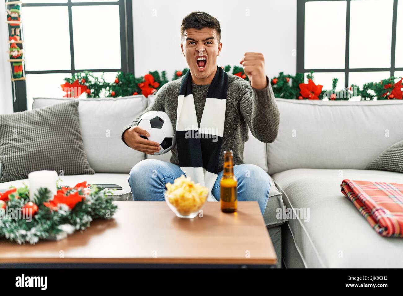 Young hispanic man football hooligan holding ball and beer annoyed and ...