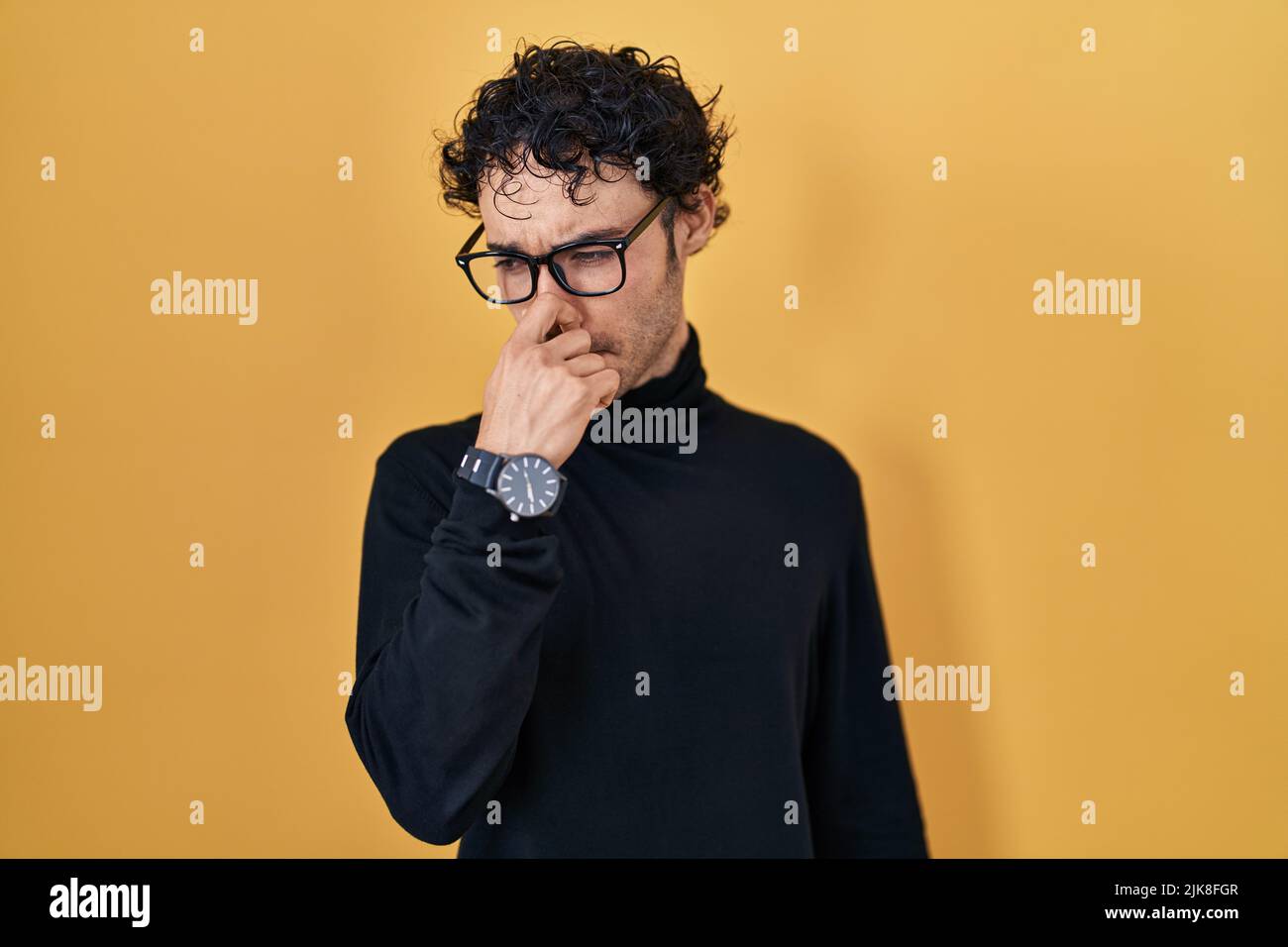 Hispanic man standing over yellow background smelling something stinky ...