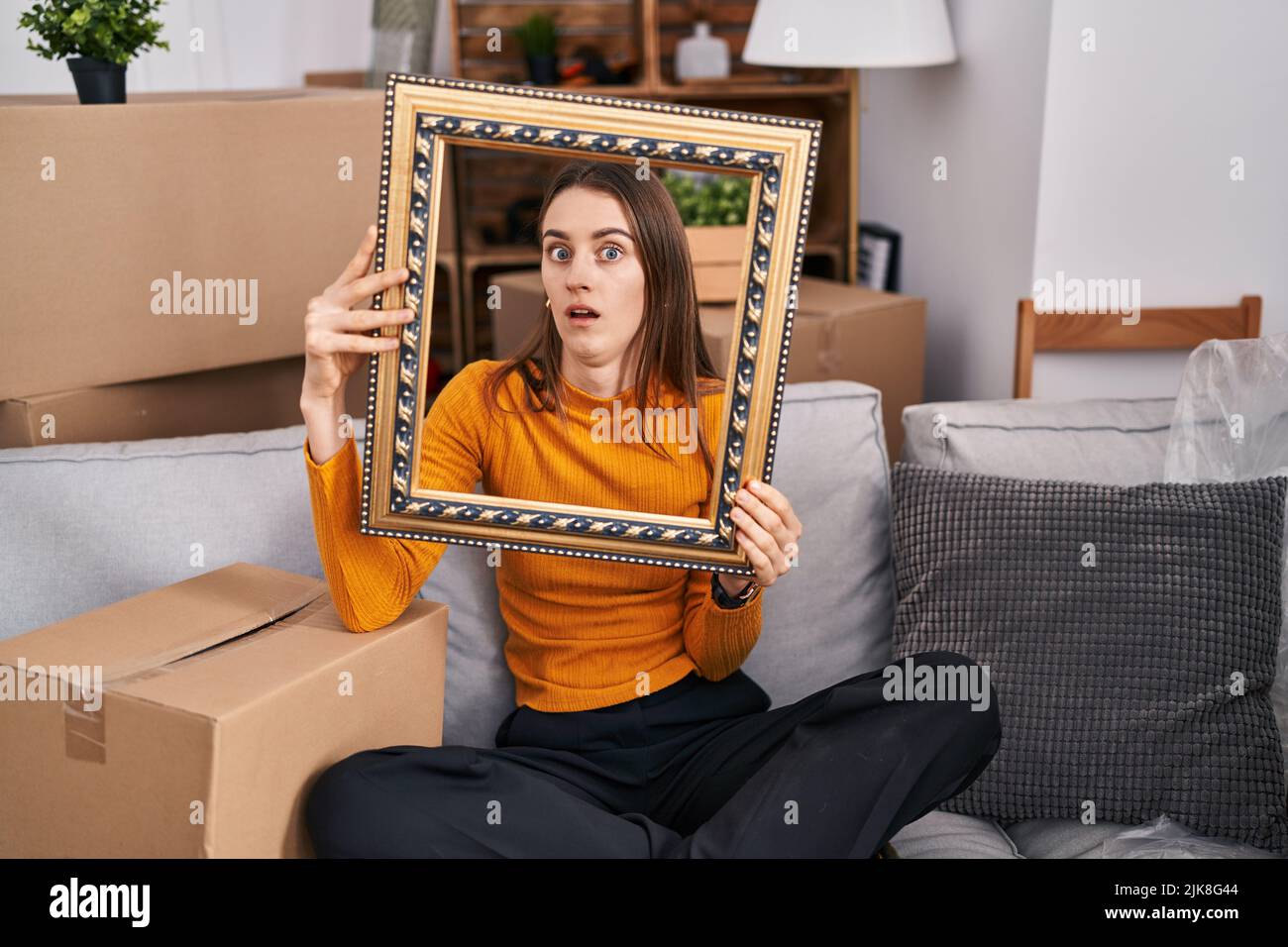 Young caucasian woman at new home holding empty frame in shock face ...