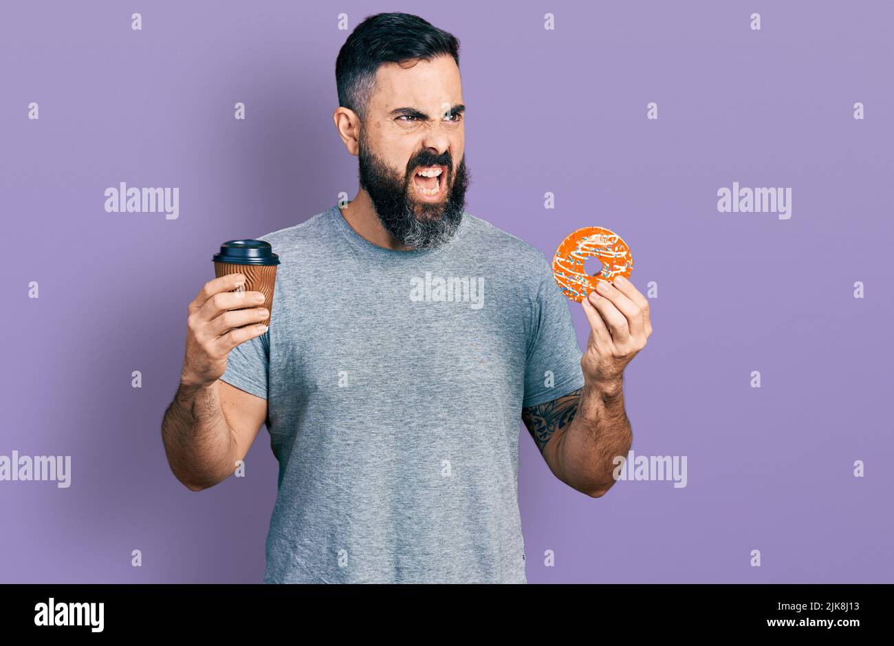 Hispanic man with beard eating doughnut and drinking coffee angry and ...