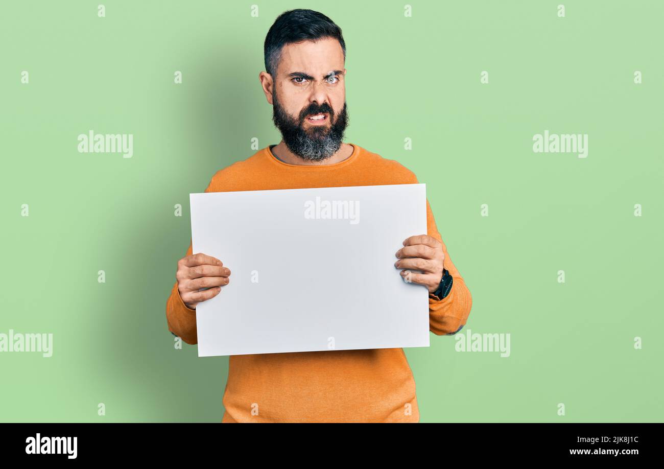 Hispanic man with beard holding blank empty banner clueless and ...