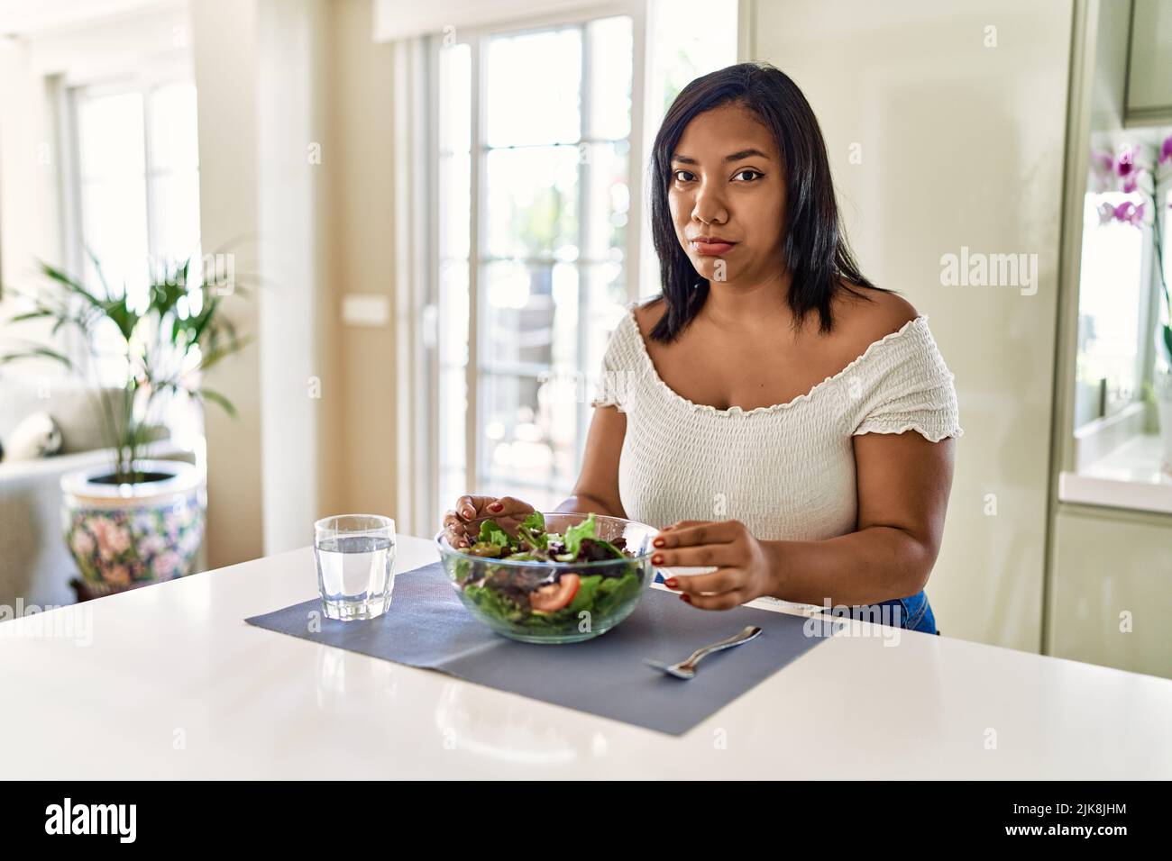 Young hispanic woman eating healthy salad at home depressed and worry ...