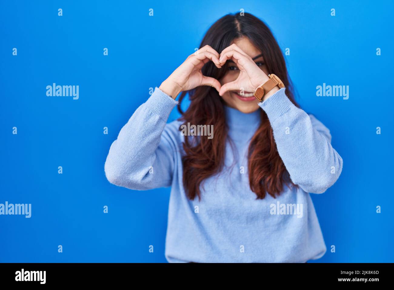 Hispanic young woman standing over blue background doing heart shape ...