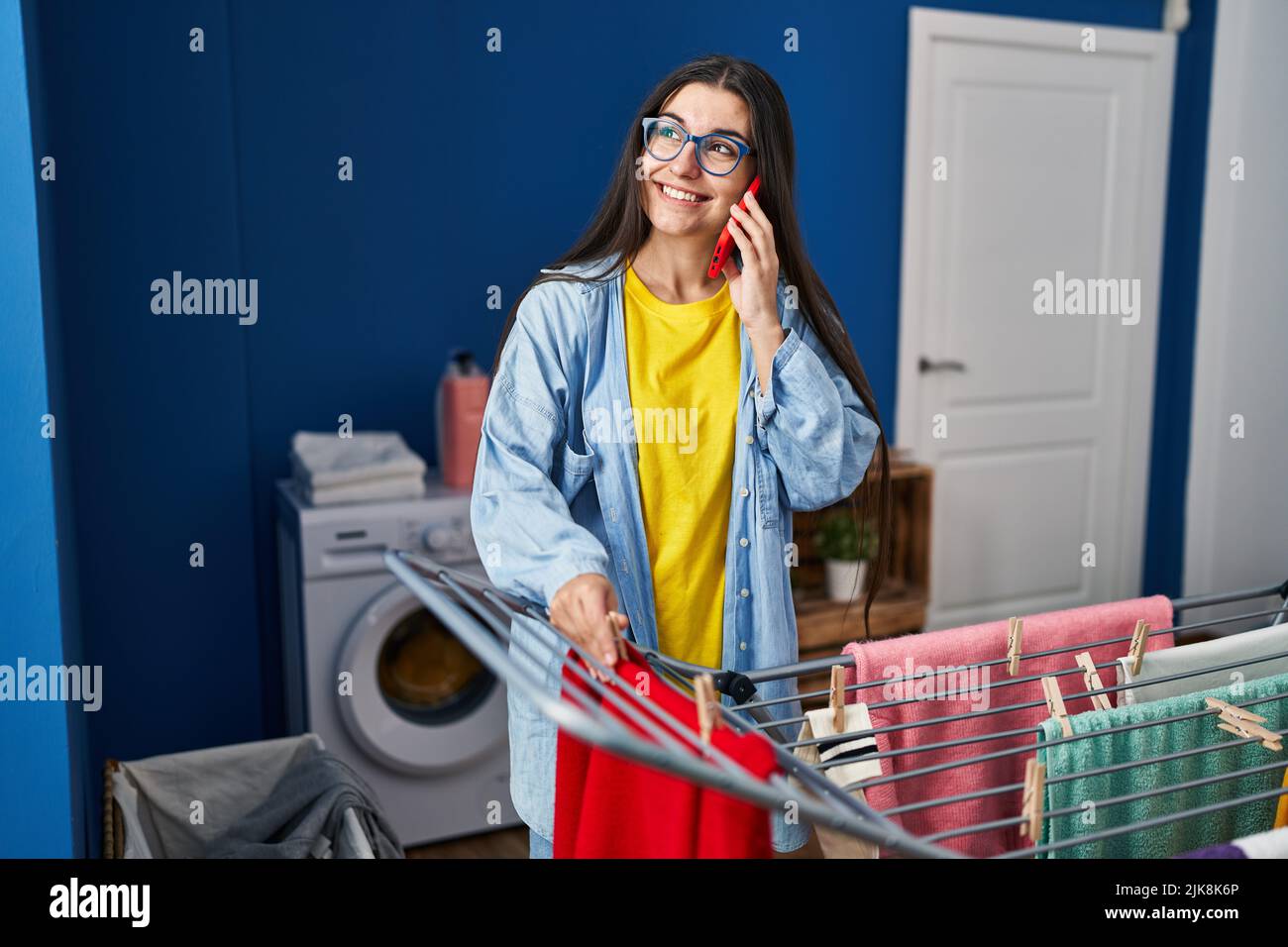 Young hispanic woman talking on the smartphone hanging clothes on ...