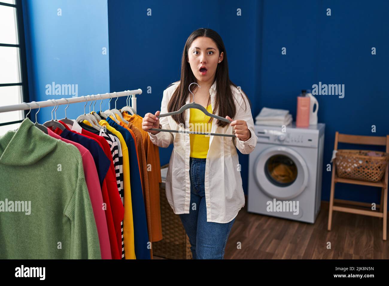 Young latin woman putting clean laundry on hangers afraid and shocked ...
