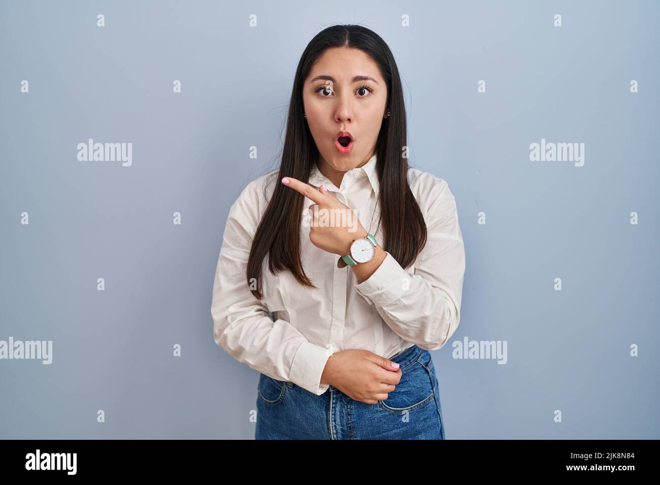 Young latin woman standing over blue background surprised pointing with ...
