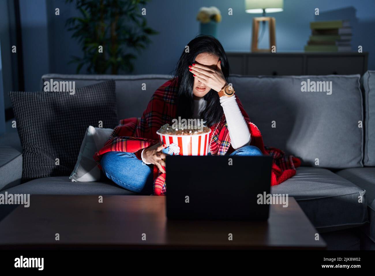 Hispanic woman eating popcorn watching a movie on the sofa covering ...