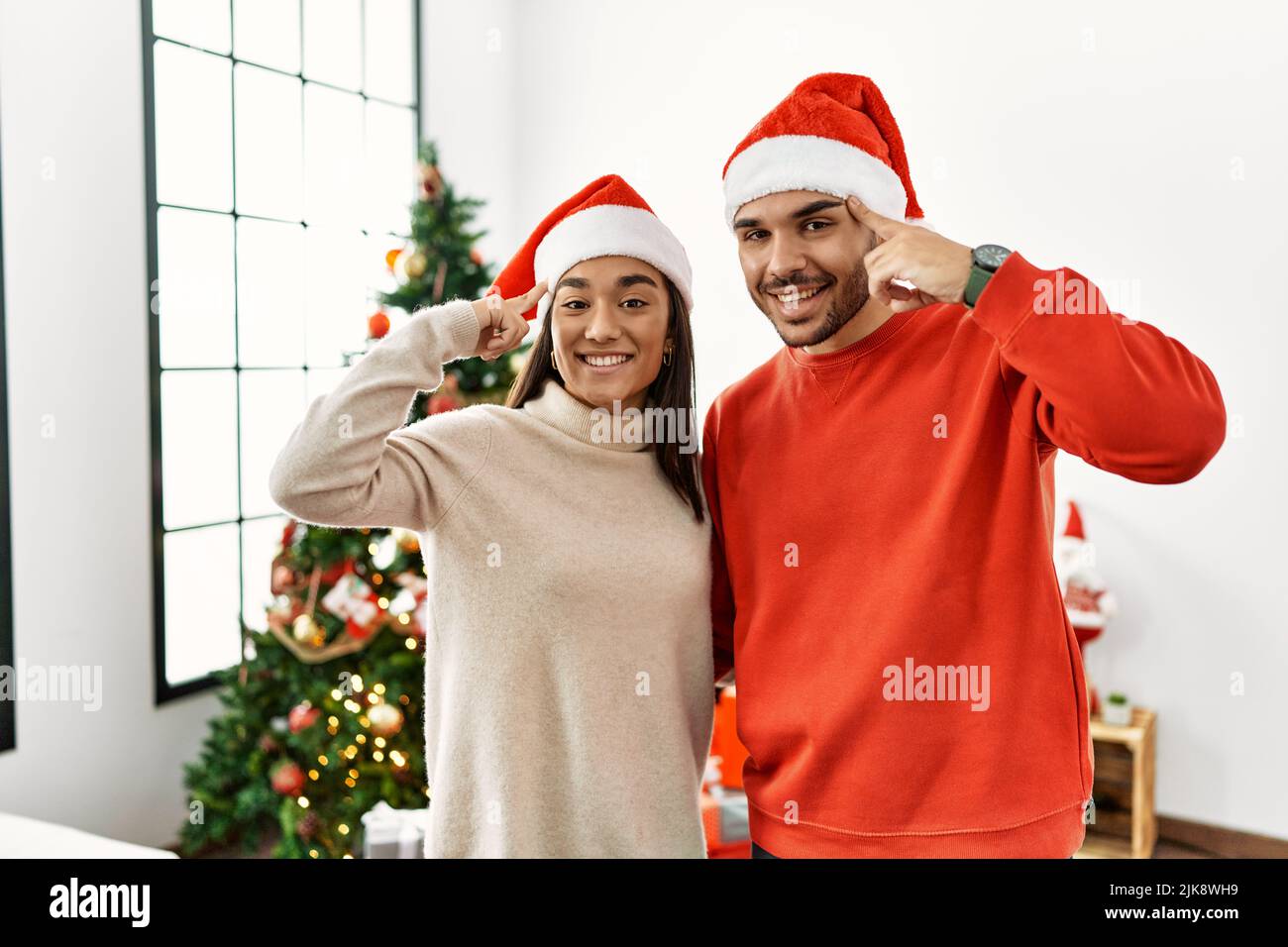 Young hispanic couple standing by christmas tree smiling pointing to ...