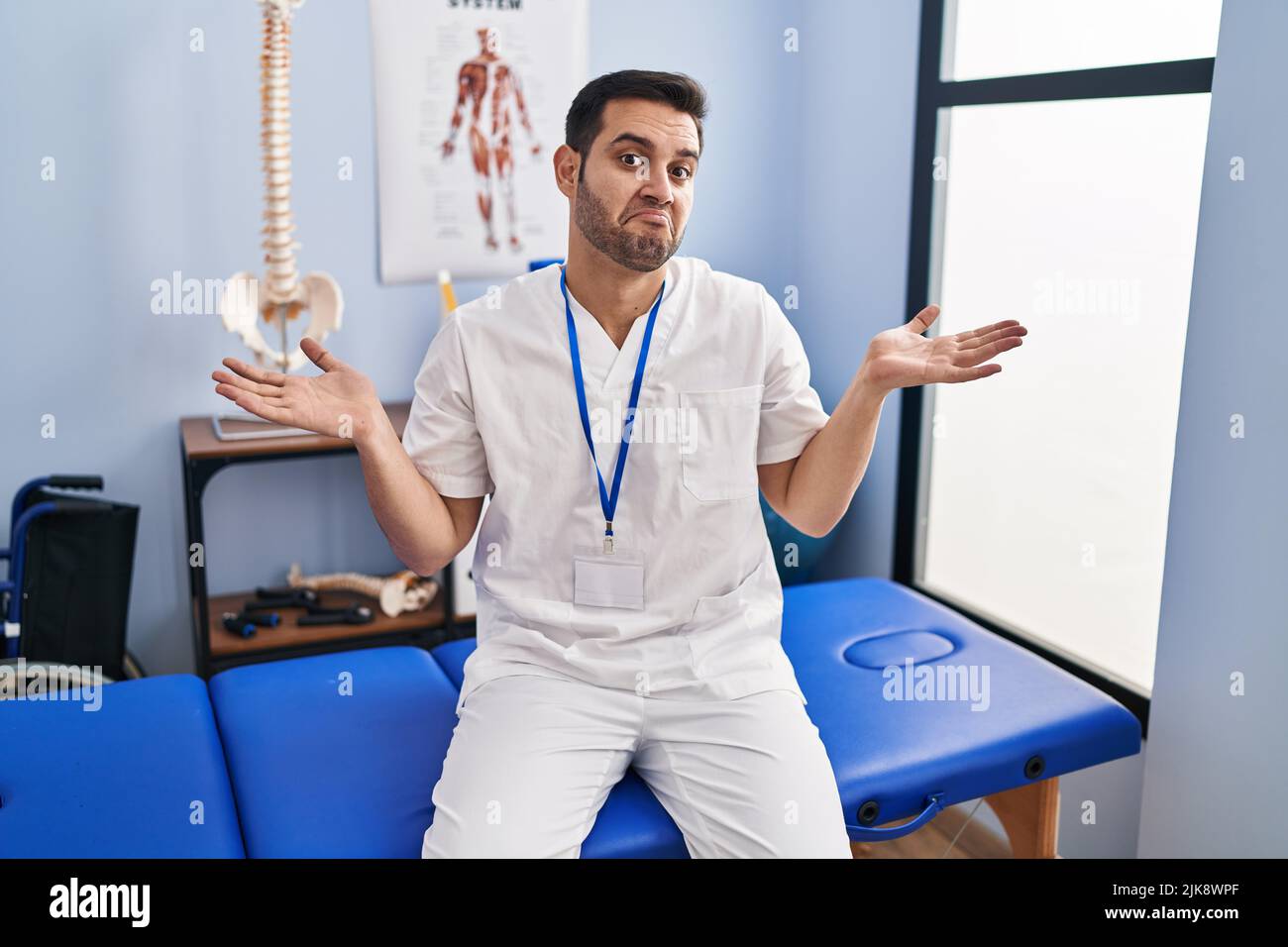 Young hispanic man with beard working at pain recovery clinic clueless ...
