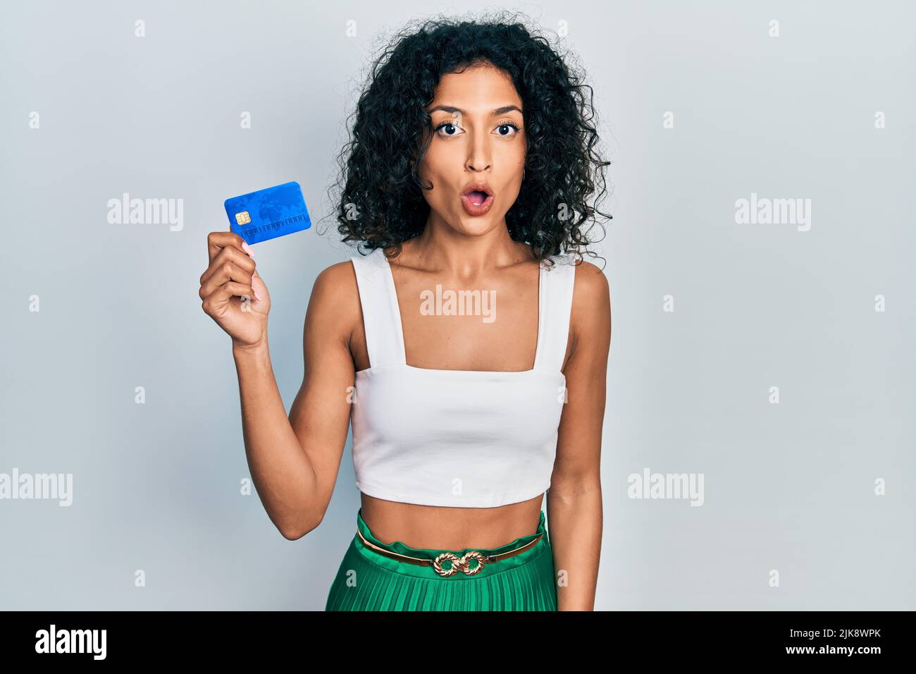 Young latin girl holding credit card scared and amazed with open mouth ...