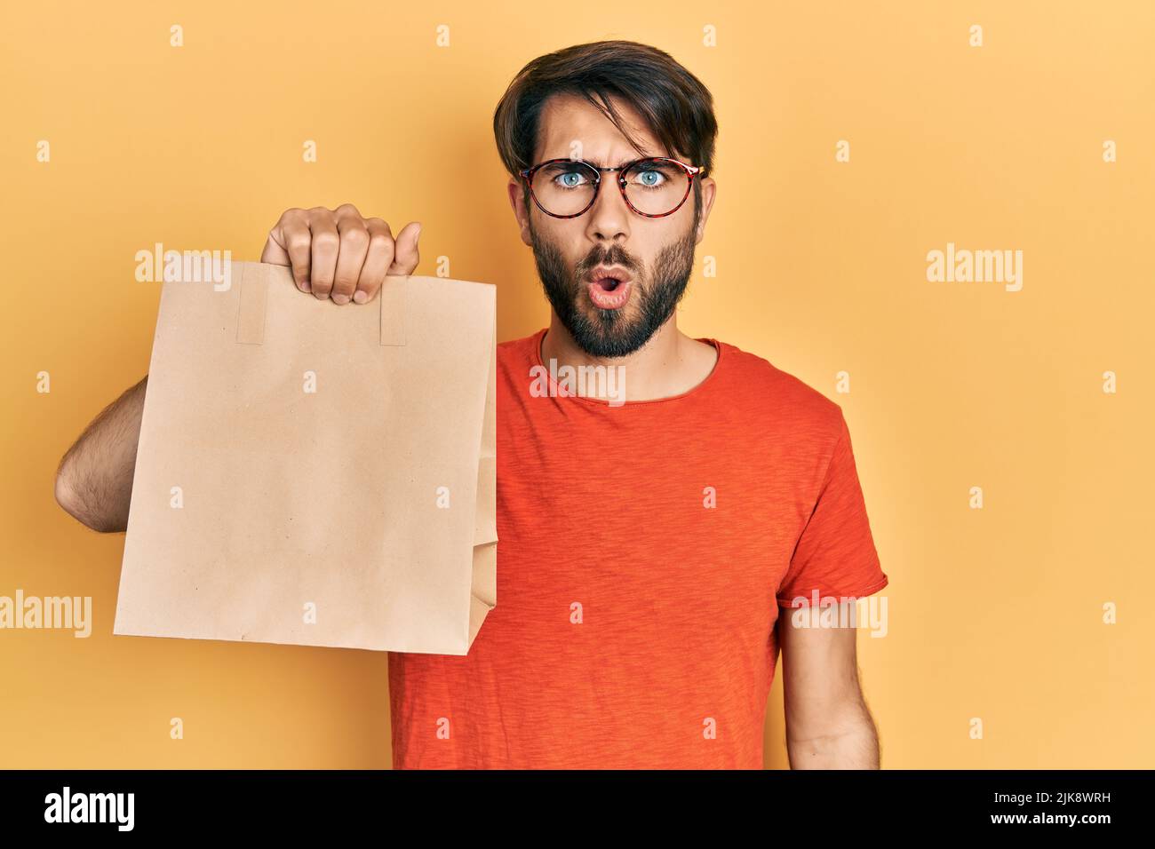 Young hispanic man holding paper bag with bread scared and amazed with ...