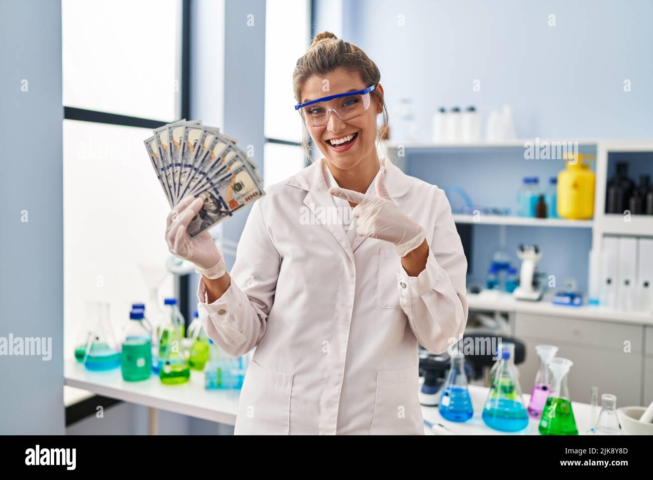 Young woman working at scientist laboratory holding money smiling happy pointing with hand and ...