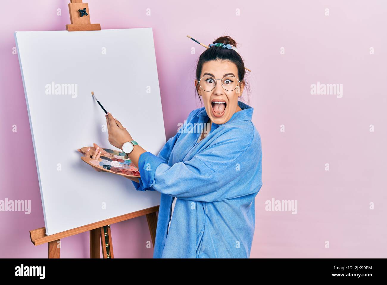 Young hispanic woman standing drawing with palette by painter easel ...