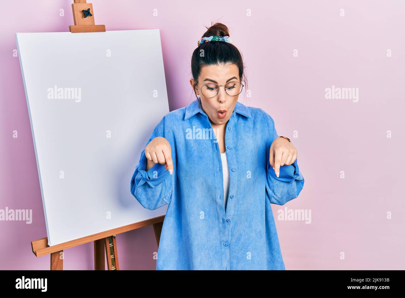 Young hispanic woman standing by painter easel stand pointing down with ...
