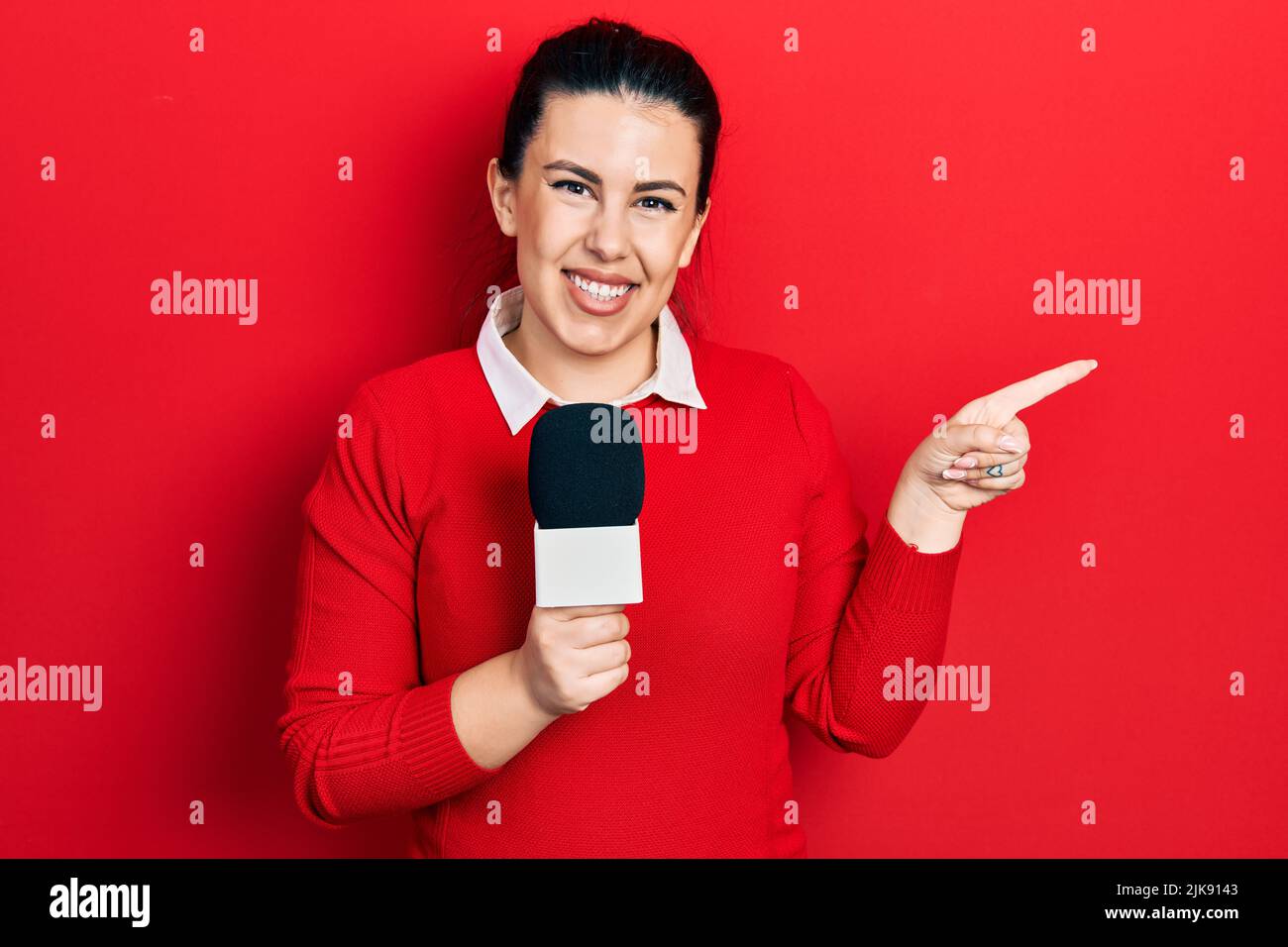 Young hispanic woman holding reporter microphone smiling happy pointing ...