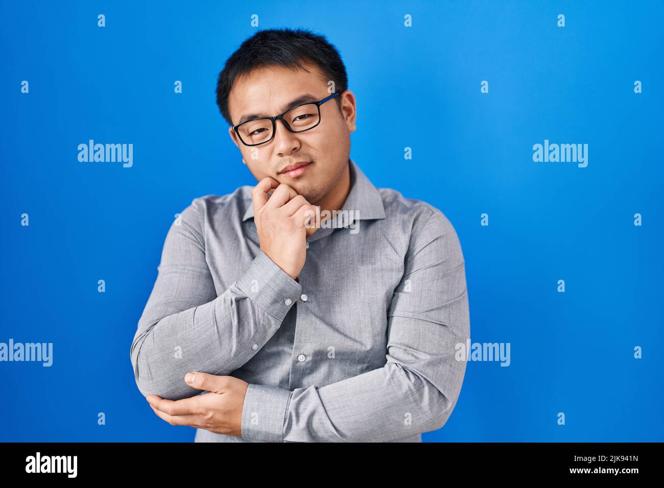 Young chinese man standing over blue background with hand on chin ...