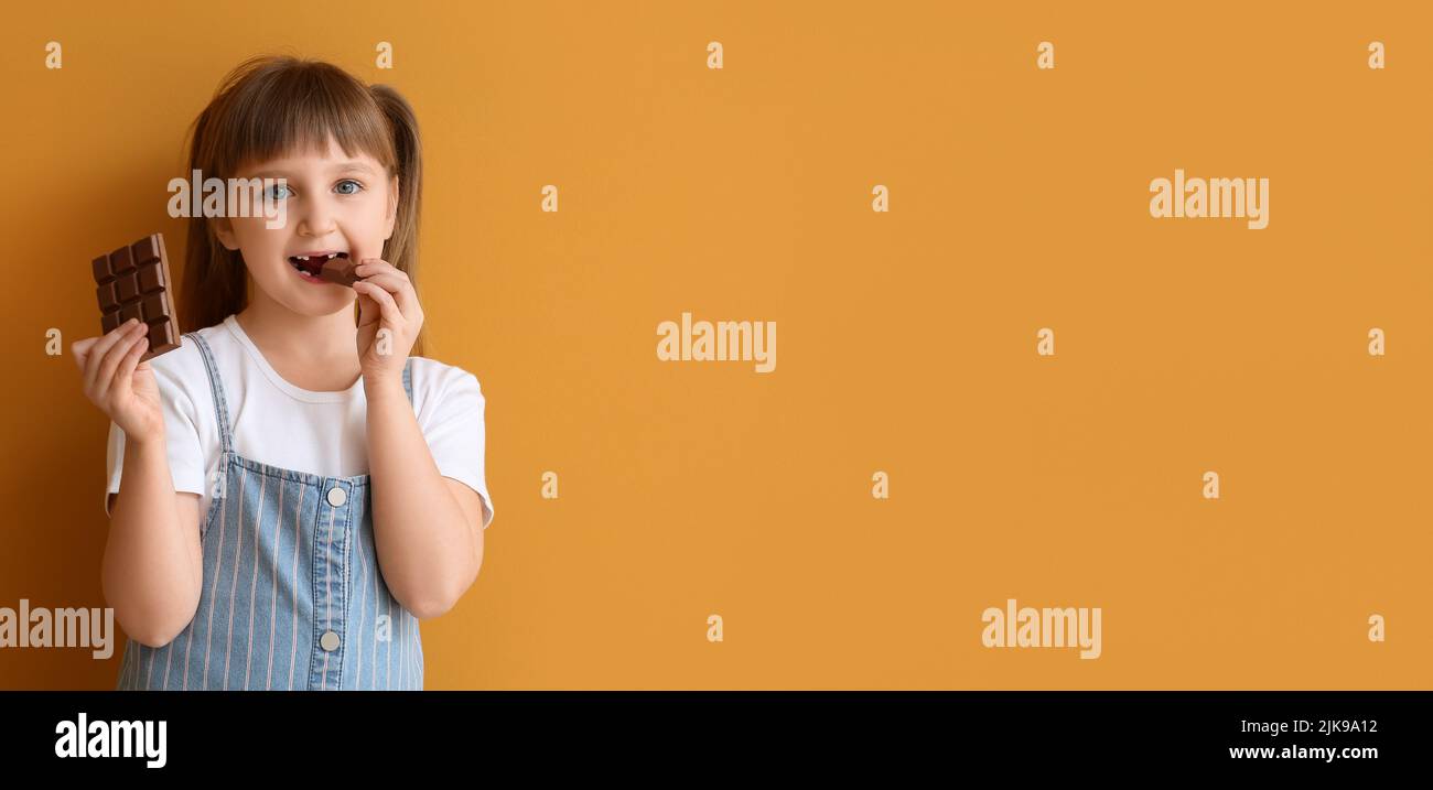 Cute little girl eating sweet chocolate on orange background with space ...