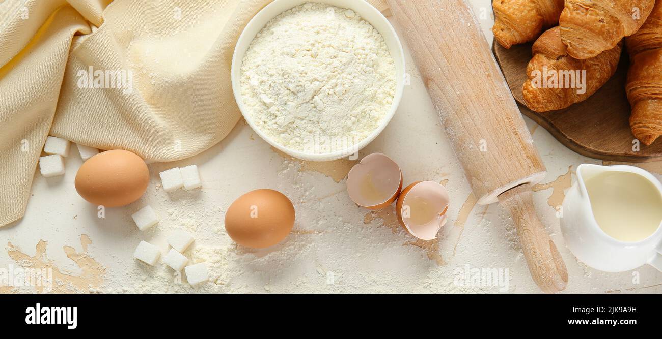 Bowl of wheat flour with raw eggs, sugar, milk, rolling pin and baked croissants on white table ...