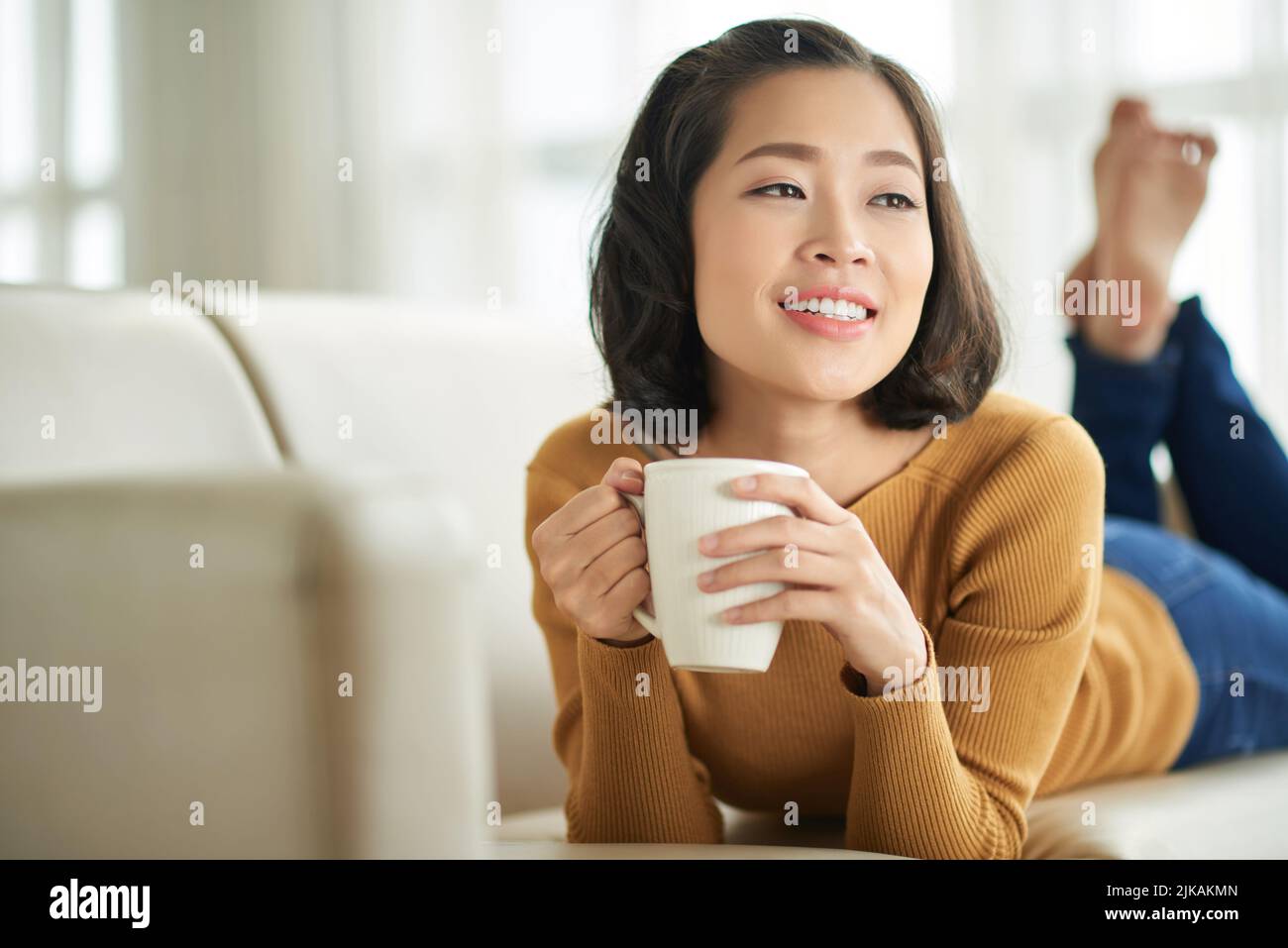Lovely Asian young woman with cup of coffee lying on sofa Stock Photo ...