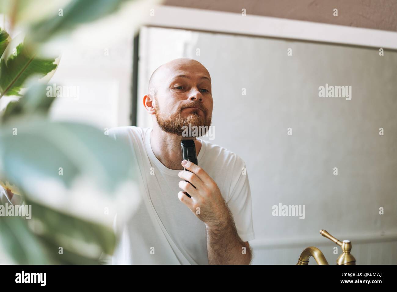 Handsome young bearded man trimming his beard with machine in bathroom ...