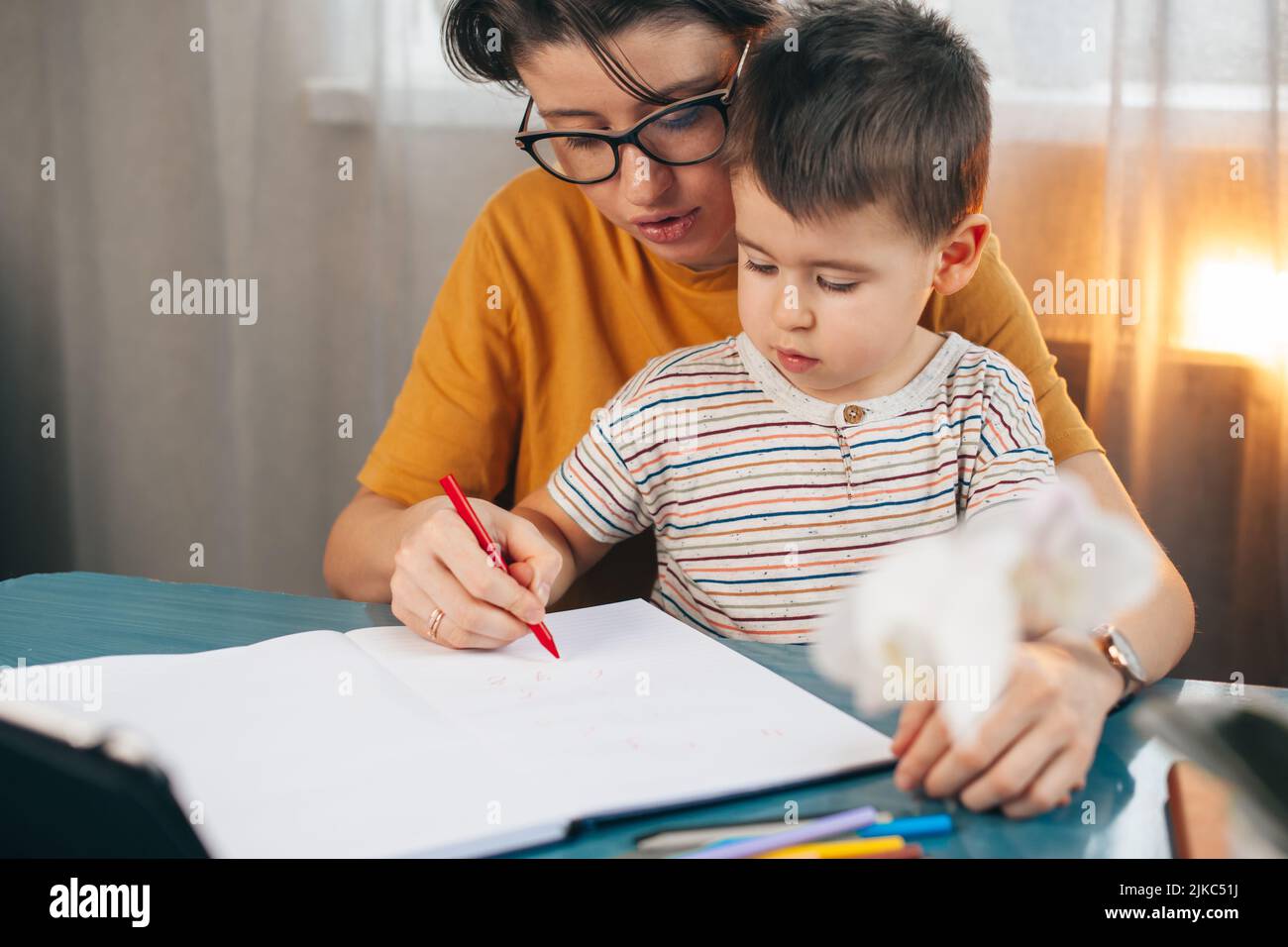 Mother teaching her small son to write while sitting on the table ...