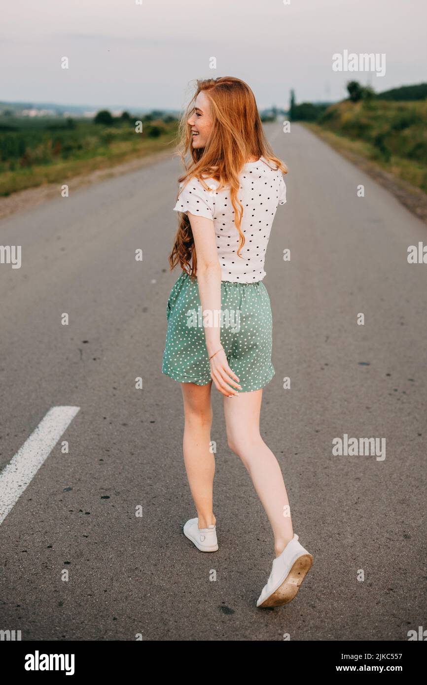 Back view of a woman walking on urban asphalt road among field outside ...