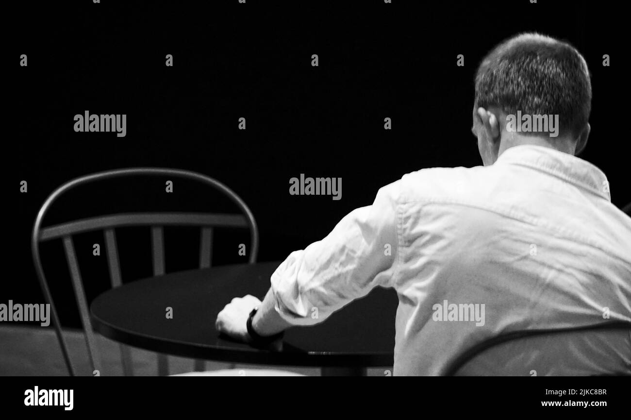 The grayscale of an adult man sitting at the table in the dark room ...