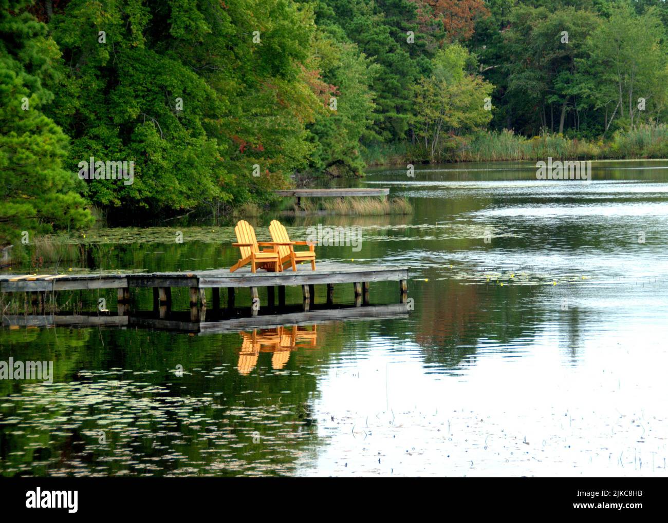 A pair of wooden chairs on a duck in the middle of a shiny lake with ...