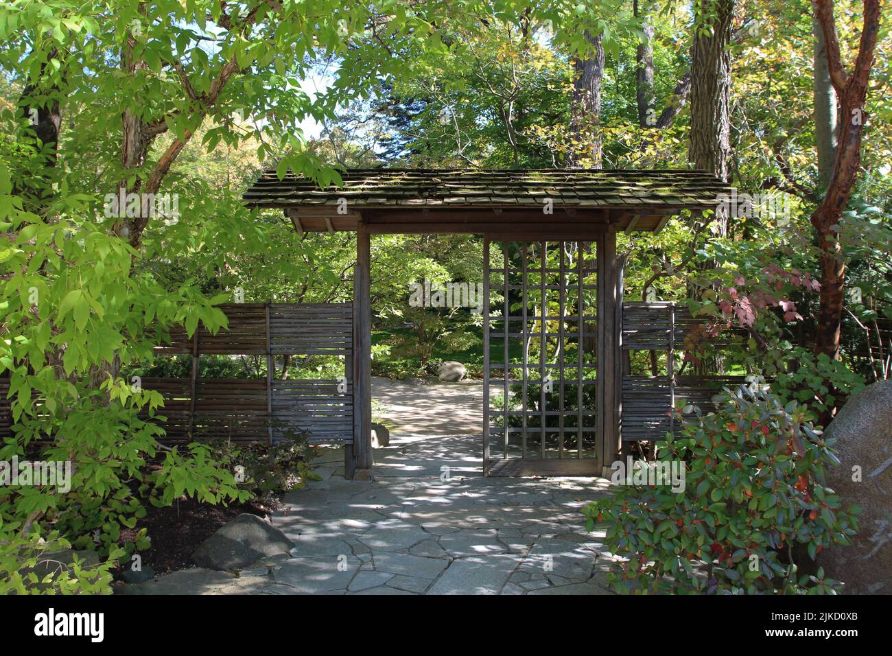 A stone path leading through a Japanese Arbor lined by shrubs and trees ...