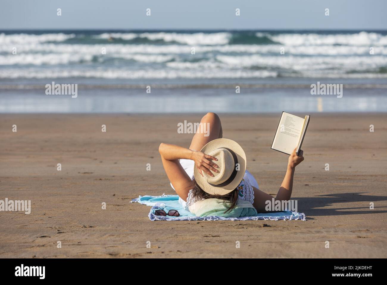 Anonymous Woman Touching Hat And Reading Book While Lying On Sand