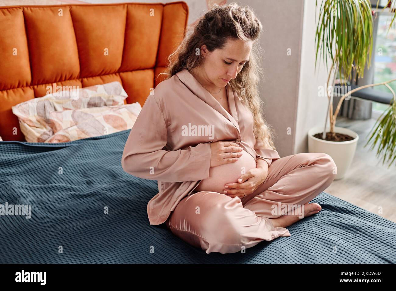 Young pregnant cross legged woman in silk pink pajamas keeping hands on ...