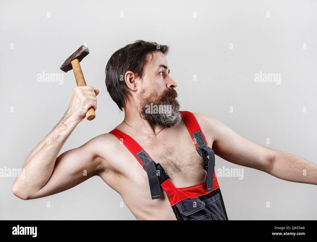 Stern bearded man in overalls swings a hammer. Builder with a hammer ...
