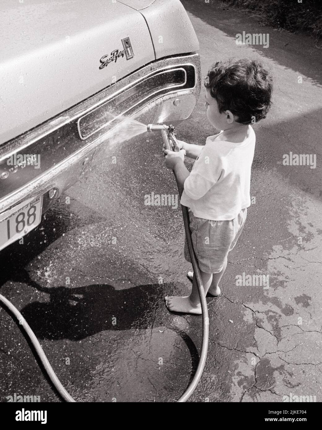 1970s YOUNG BOY HELPING WITH WASHING THE CAR STANDING IN HIS BARE FEET ...