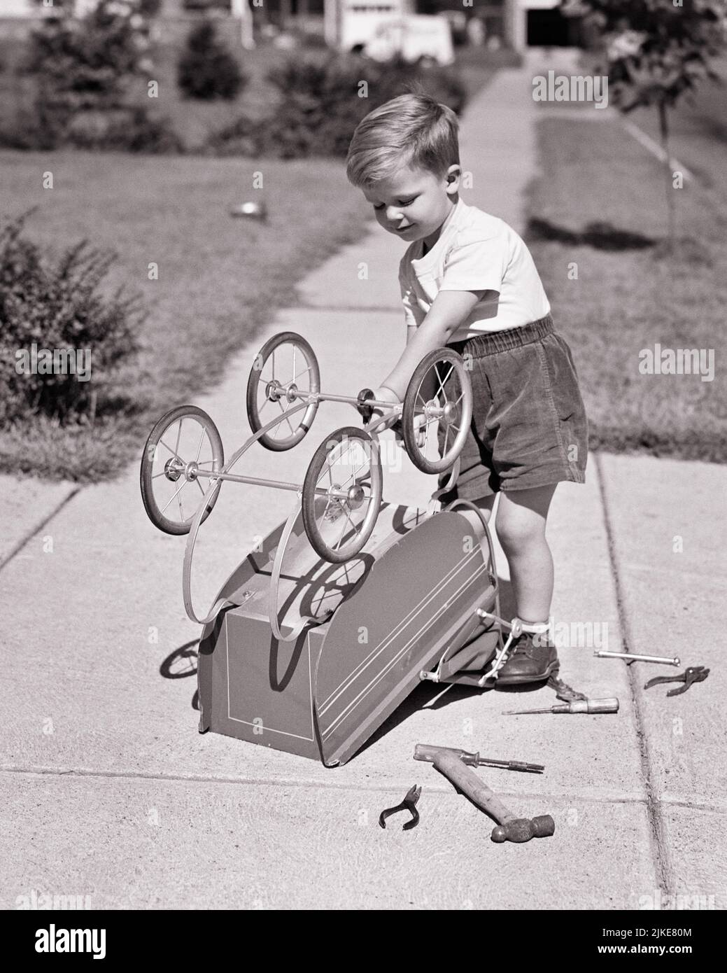 1940s LITTLE BOY REPAIRING TOY BABY CARRIAGE ON SUBURBAN SIDEWALK USING ...