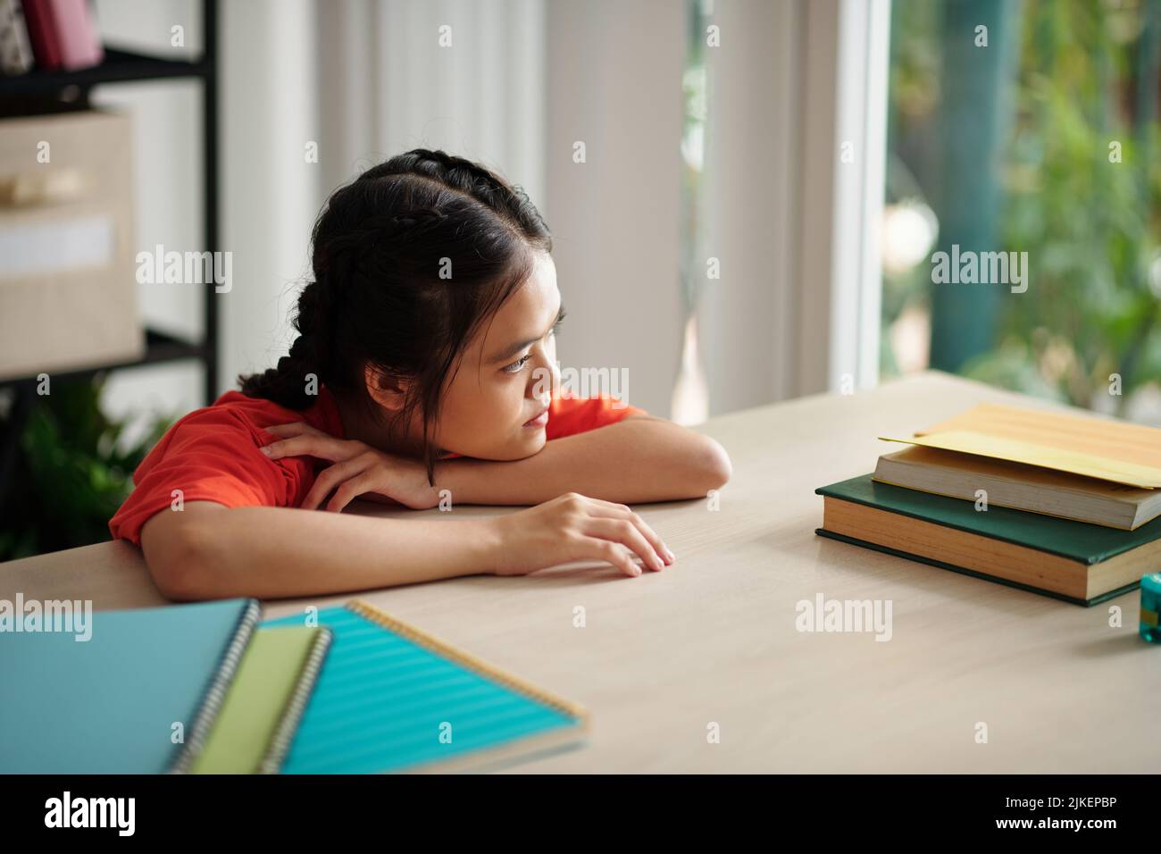 Pensive Asasian schoolgirl leaning on desk in class and looking through ...