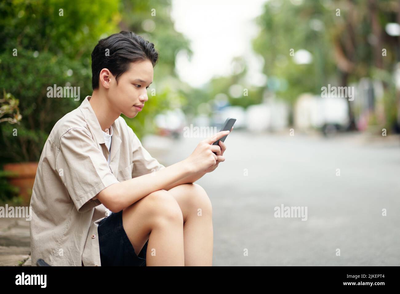 Teenage boy with smartphone sitting on pavement and texting friends ...