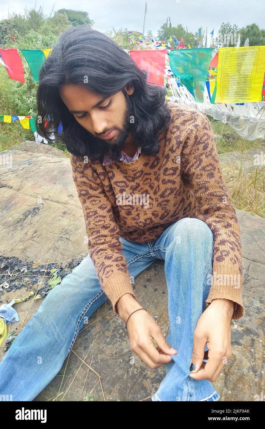 A young Indian male tourist with long hair and beard sitting over rock ...