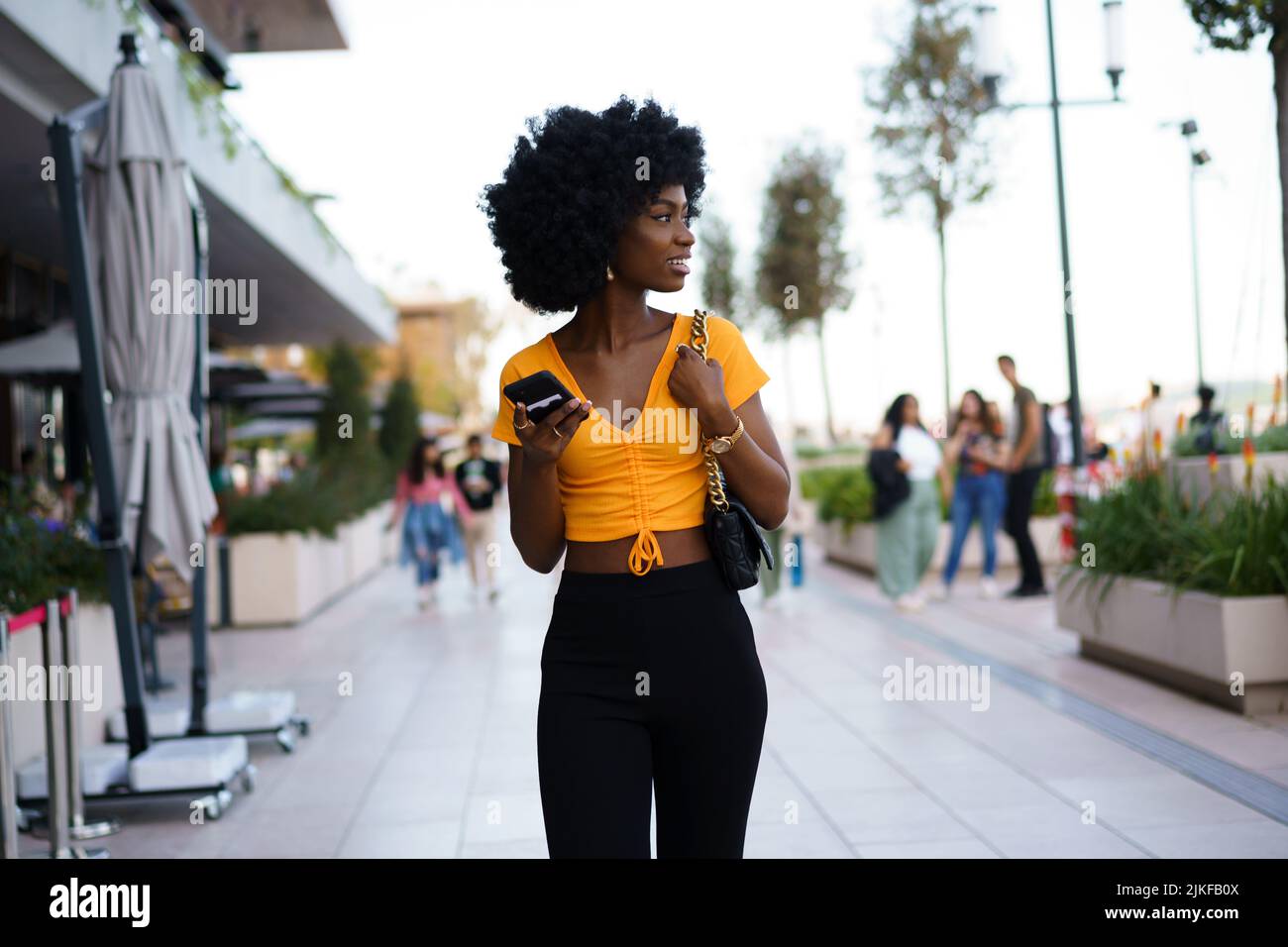 Smiling afro-american woman holding mobile phone while walking in the ...