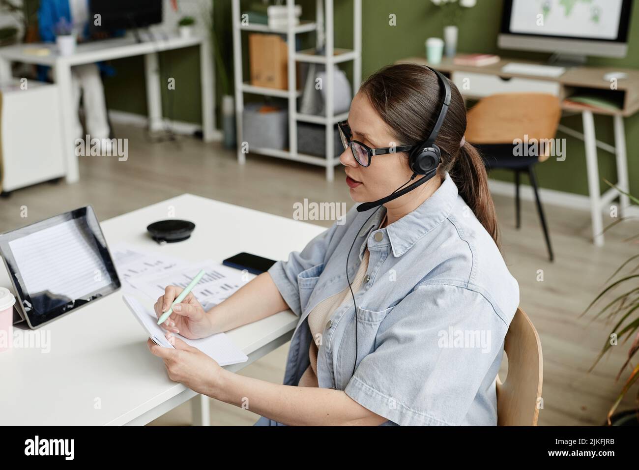 Young businesswoman in headphones making notes while talking to ...