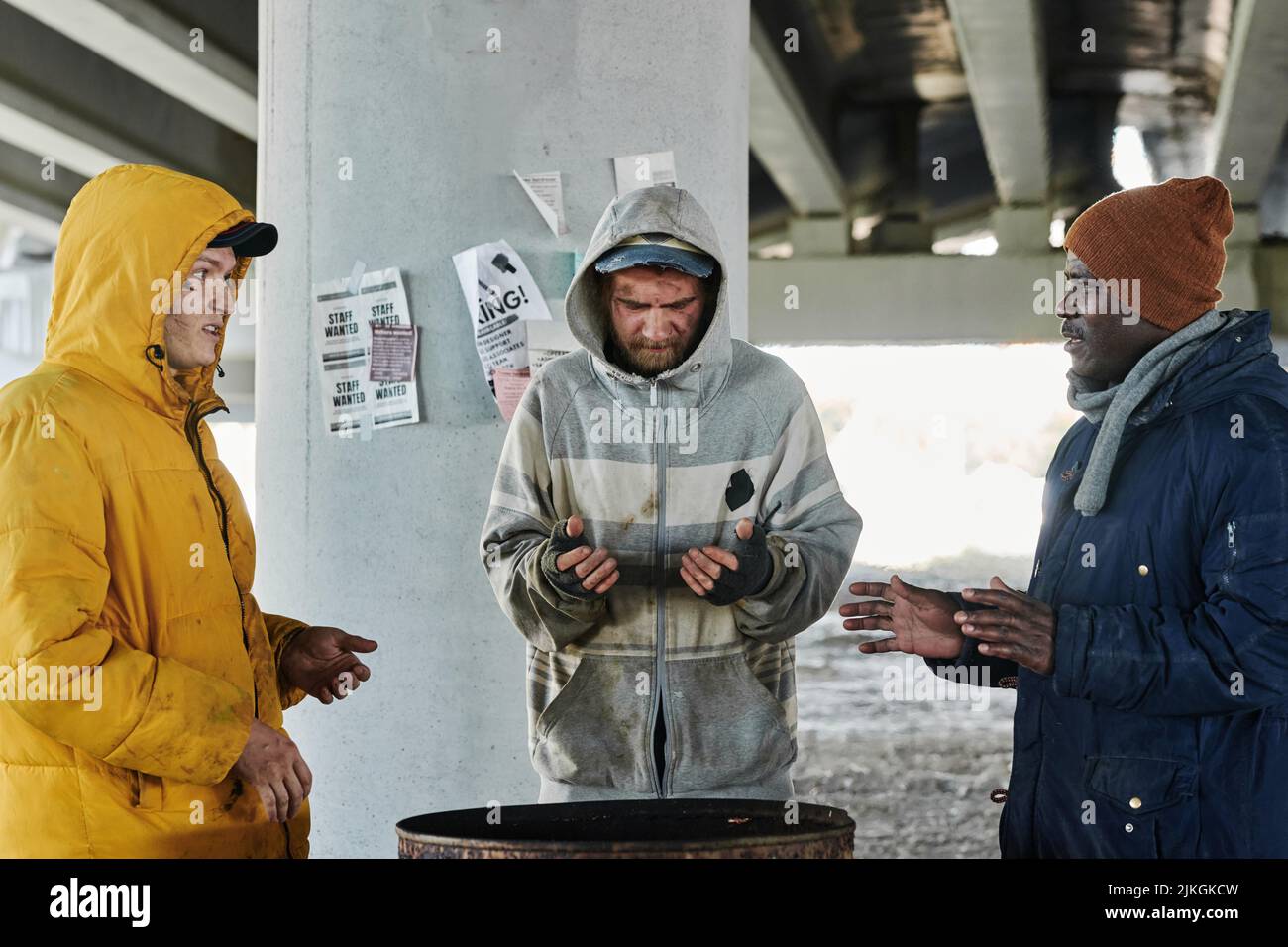 Homeless people in torn and dirty clothes standing around barrel of ...