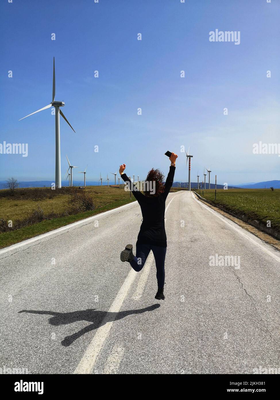 A vertical shot of a female happily jumping on a road through a field ...