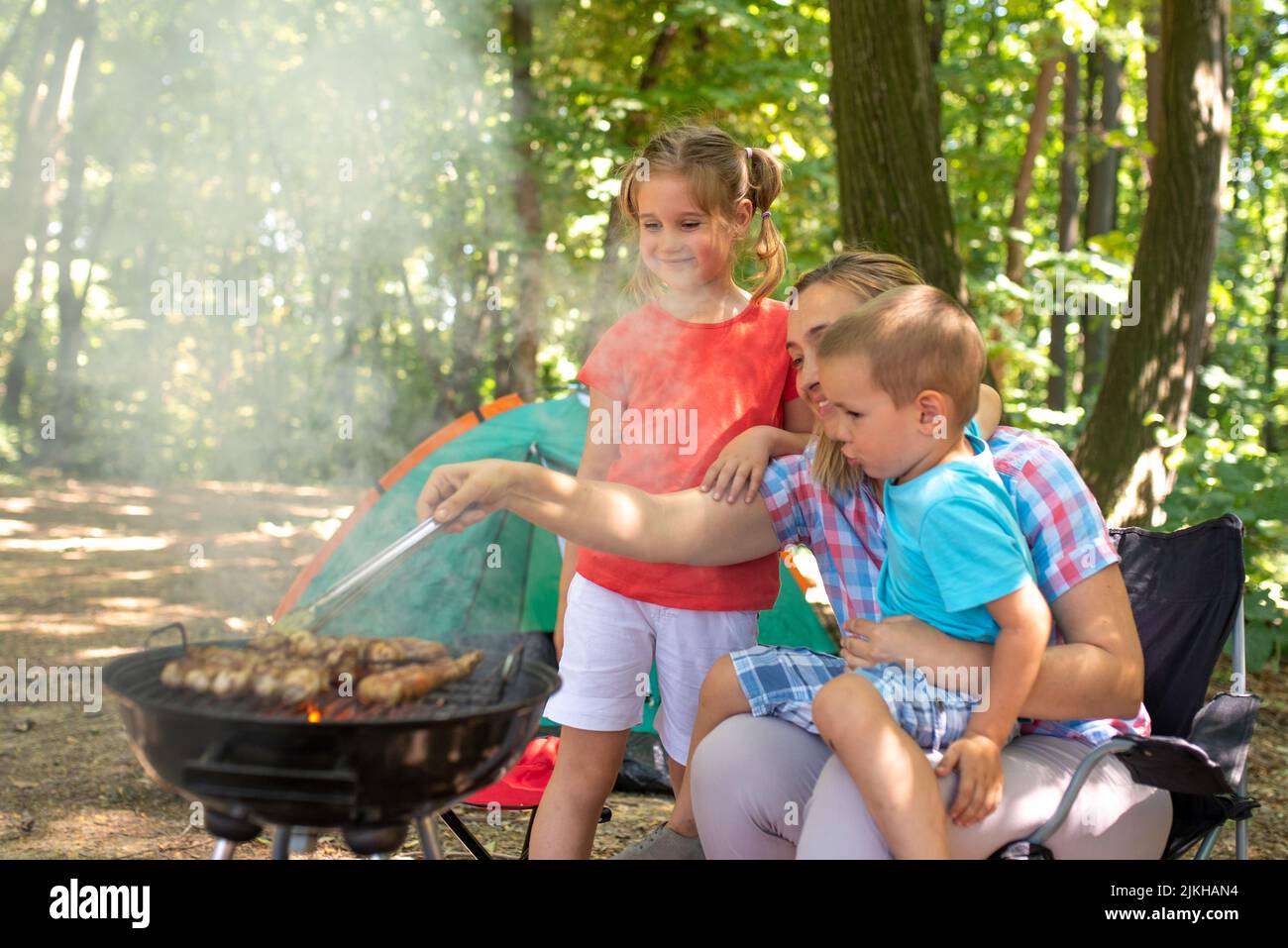 A young happy mother camping and cooking barbecue in a forest with her ...