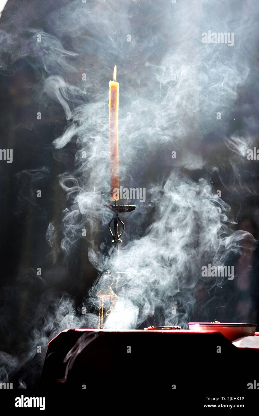 A vertical shot of a lit candle in a stand with burned incense smoke in ...