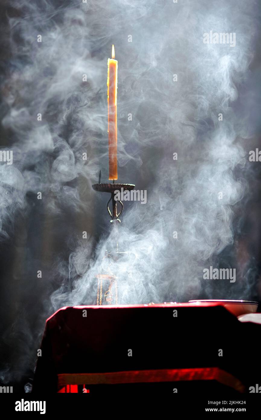 A vertical shot of a lit candle in a stand with burned incense smoke in ...