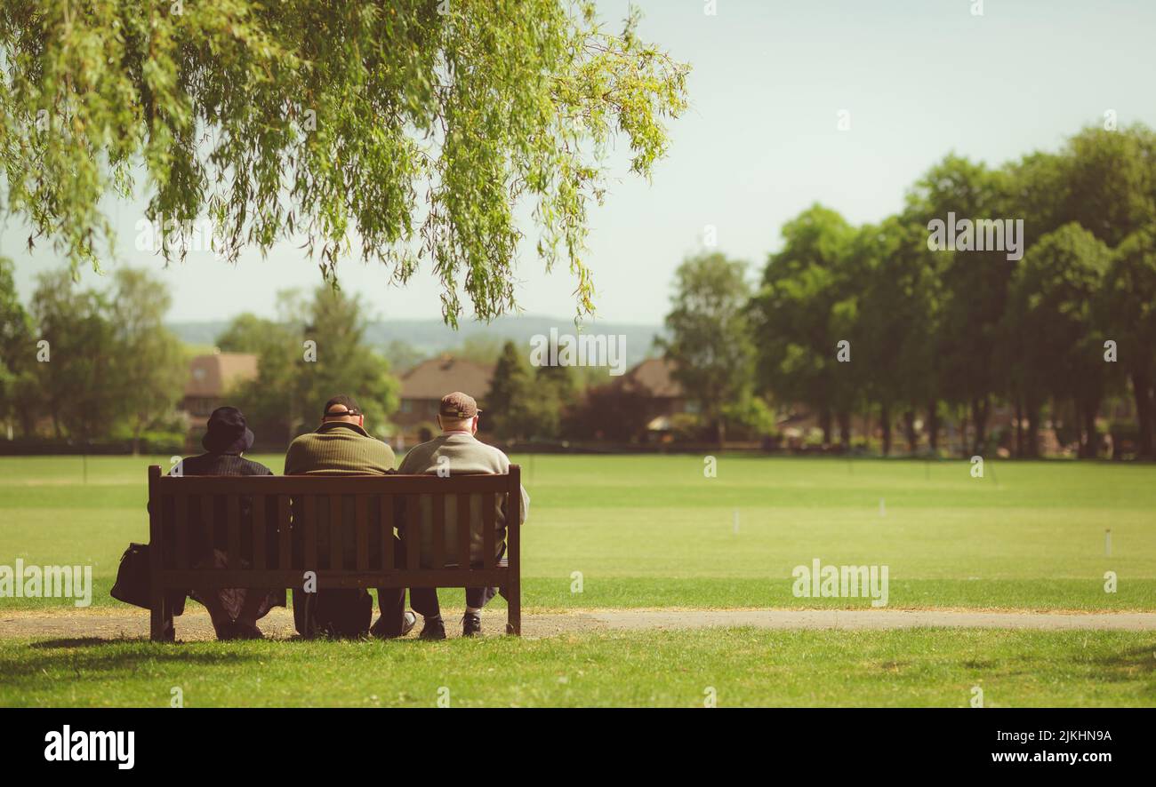 A bright summer day outdoors with three people sat on a bench in a park ...
