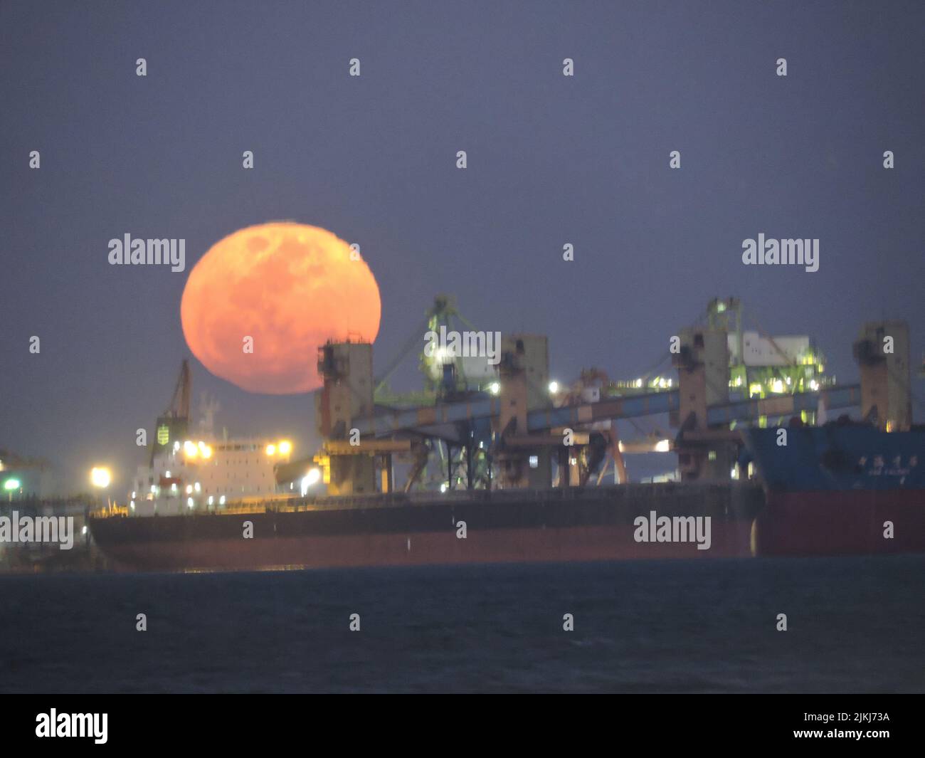 A shot of a bright yellow supermoon with a foreground of a cargo ship ...