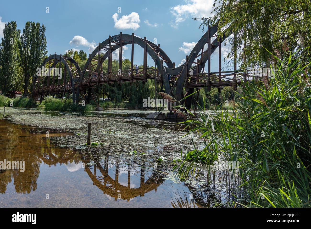 A view of an arch bridge with people passing over the small muddy lake ...