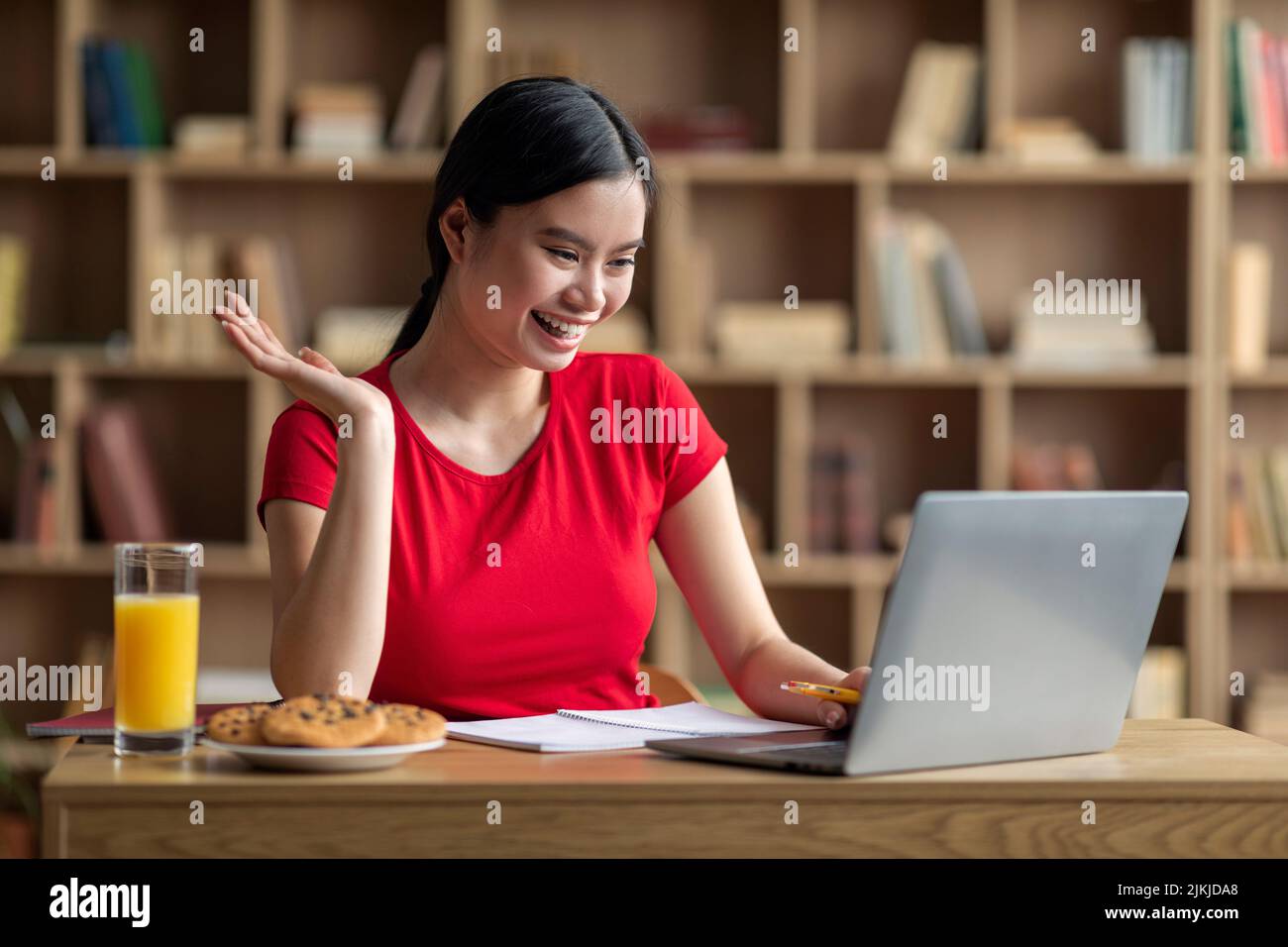 Glad teenager japanese lady student say hello, waving hand, watching online lesson in room ...