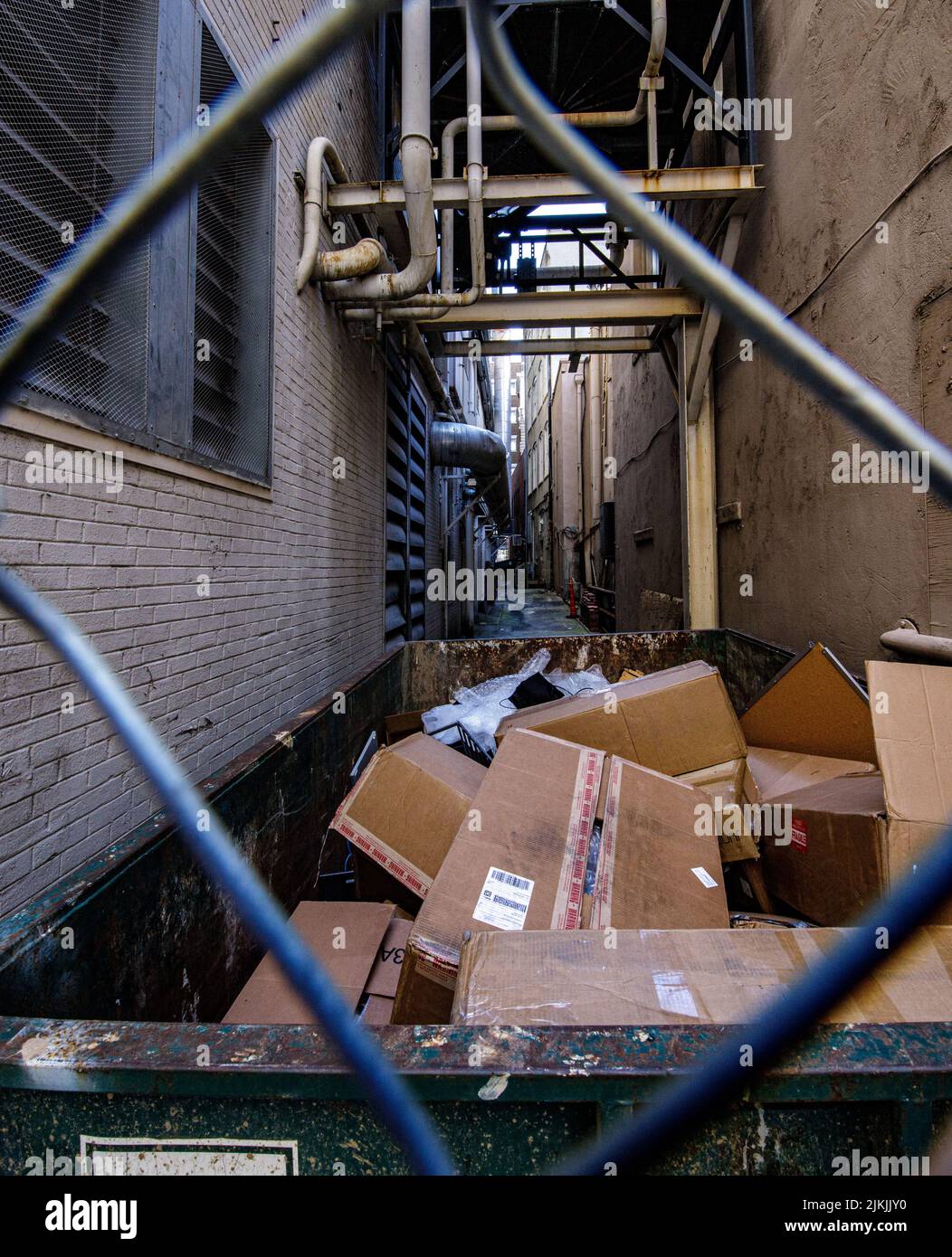 A pile of boxes on a narrow space between buildings with a chain-link ...